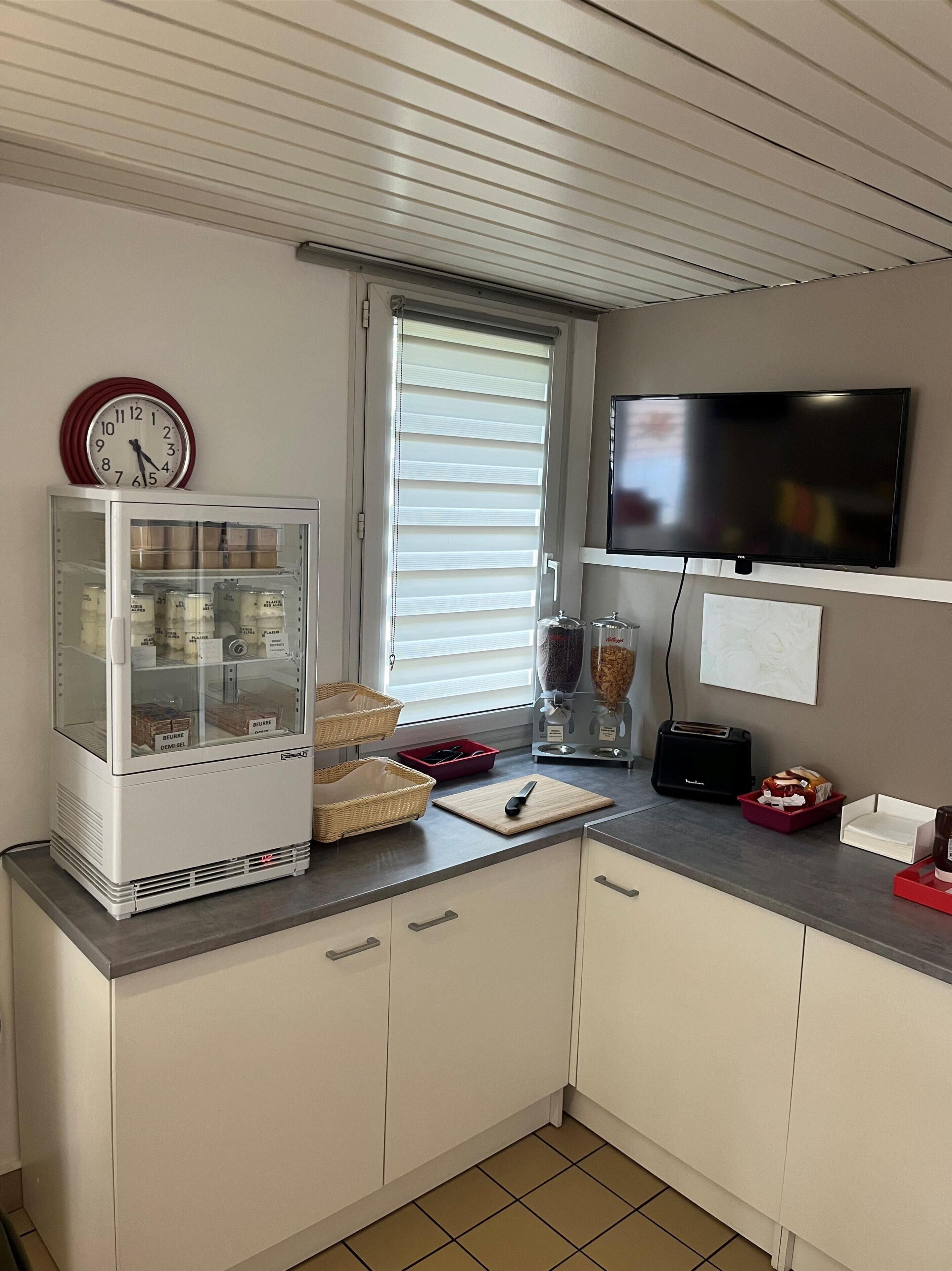 Uma cozinha compacta com geladeira, torradeira, relógio, janela com persianas e televisão na parede.
