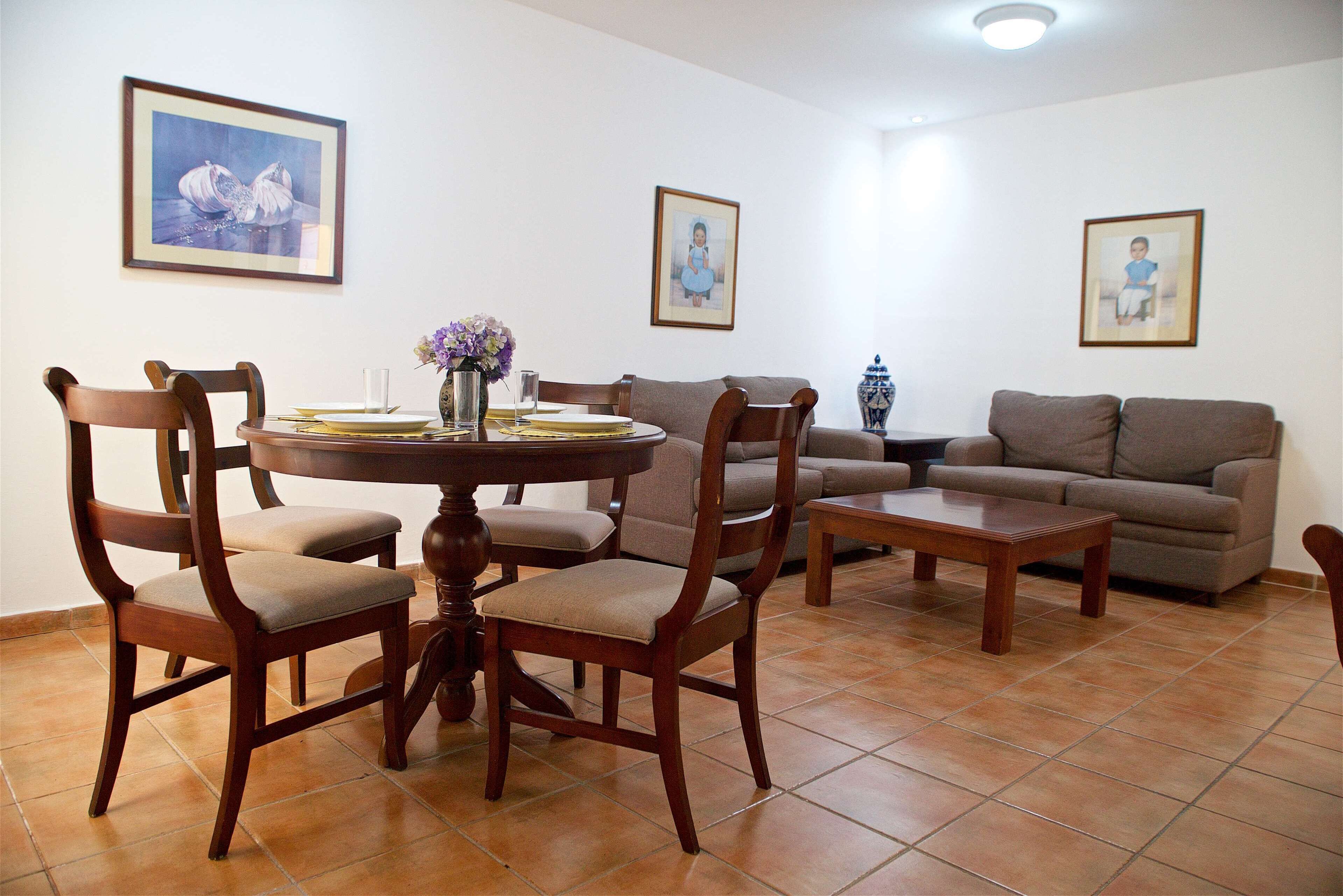 Uma área de refeições com uma mesa redonda, cadeiras e um vaso de flores. Há um sofá e uma mesa de centro ao fundo.