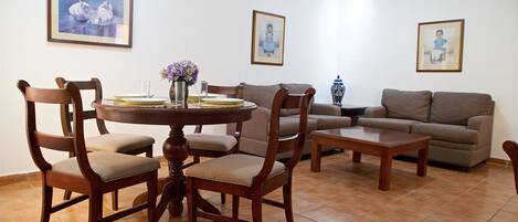 Uma área de refeições com uma mesa redonda, cadeiras e um vaso de flores. Há um sofá e uma mesa de centro ao fundo.