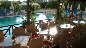 Área da piscina com espreguiçadeiras e mesas, piscina e vegetação ao fundo.