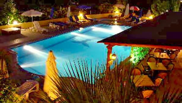 Piscina com espreguiçadeiras e guarda-sóis, cercada por edifícios com iluminação externa.