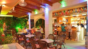 Restaurante ao ar livre com mesas e cadeiras de madeira, área de bar com diversos drinques e uma parede verde com plantas.