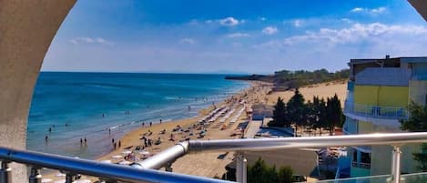 Vista da praia a partir de uma varanda com guarda-corpo, guarda-sóis e céu claro.