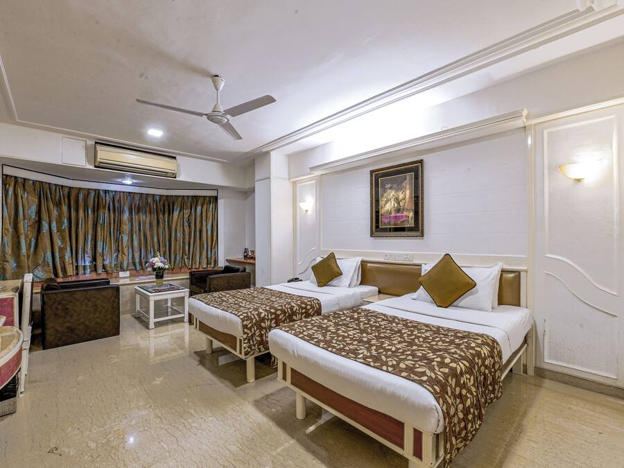 Quarto de hotel com duas camas, ventoinha no teto, ar condicionado, escrivaninha com cadeira, mesa pequena com um vaso, e cortina.