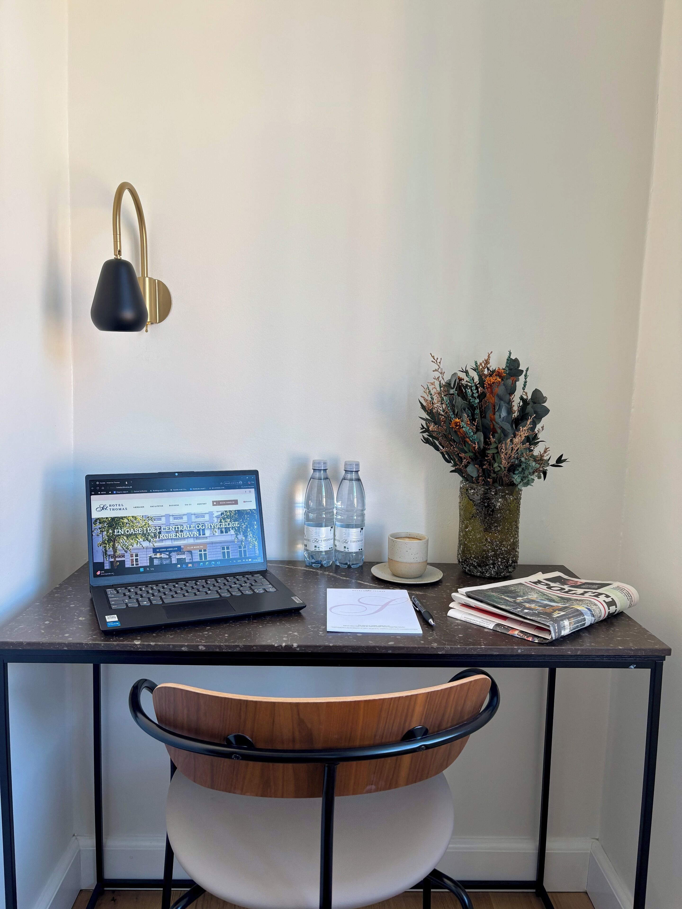 Um laptop sobre uma mesa com um vaso de plantas, duas garrafas de água, uma caneca e um jornal.