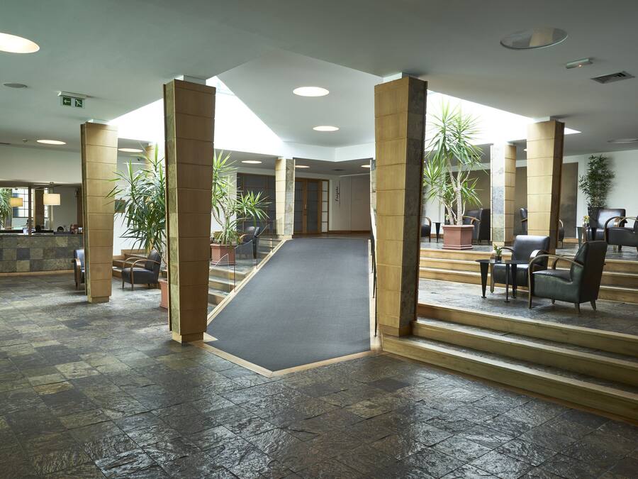 Um hall amplo com escadaria central, piso de pedra e vários vasos de plantas.