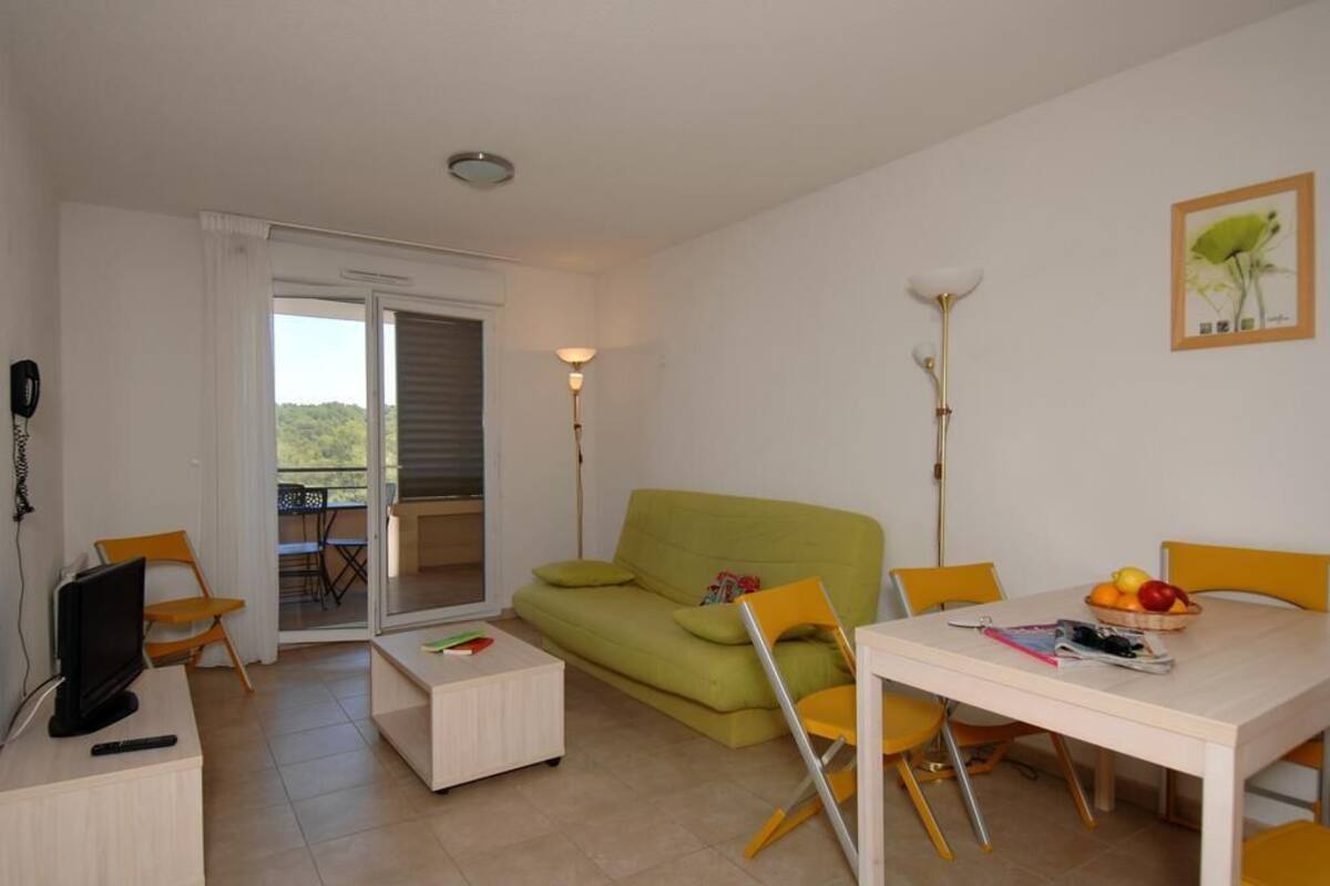 Sala de estar com sofá verde, mesa de centro branca, mesa de jantar com cadeiras amarelas, televisão e varanda com vista.