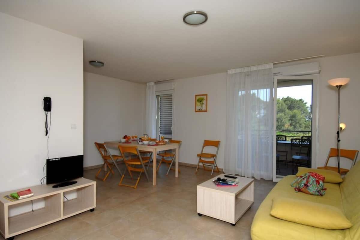 Uma sala de estar com televisão, sofá, mesa de jantar com cadeiras e varanda com vista.