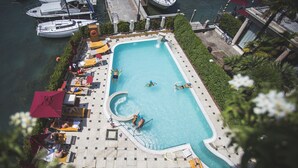 Vista aérea de uma piscina com pessoas se banhando e tomando sol, cercada por espreguiçadeiras e guarda-sóis, e barcos atracados por perto.