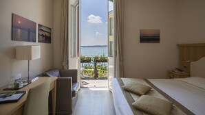 Quarto de hotel com cama, escrivaninha, cadeira, mesa de cabeceira e vista para o mar por uma janela aberta.