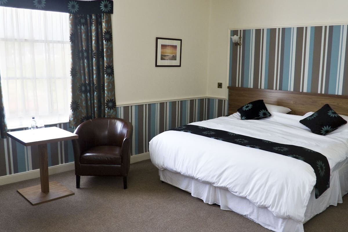 Quarto de hotel com cama, cadeira, escrivaninha e janela com cortinas.