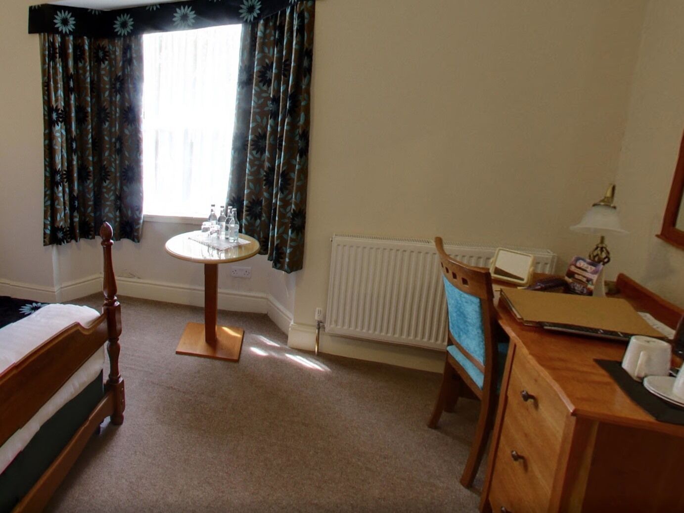 Quarto de hotel com cama, uma mesa redonda com cadeira, escrivaninha com cadeira, radiador e janela com cortinas.