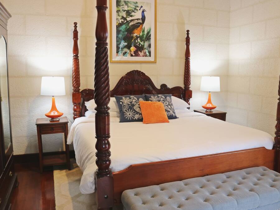 Uma cama de dossel com um banquinho estofado na frente, duas mesas de cabeceira com abajures e um quadro com a imagem de um pavão na parede.