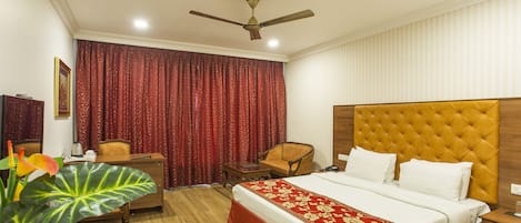 Quarto de hotel com cama, escrivaninha, cadeira, ventoinha no teto e uma planta grande.