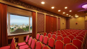 Uma sala de conferências com fileiras de cadeiras vermelhas, uma janela grande com vista para a cidade, cortinas estampadas e um lustre.