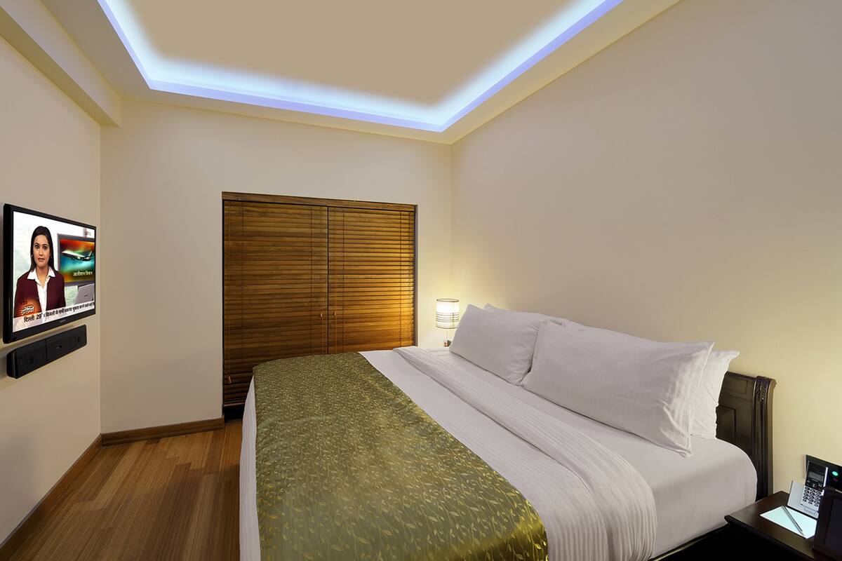 Quarto de hotel com cama, televisão na parede, cabeceira de madeira e mesa de cabeceira com abajur.