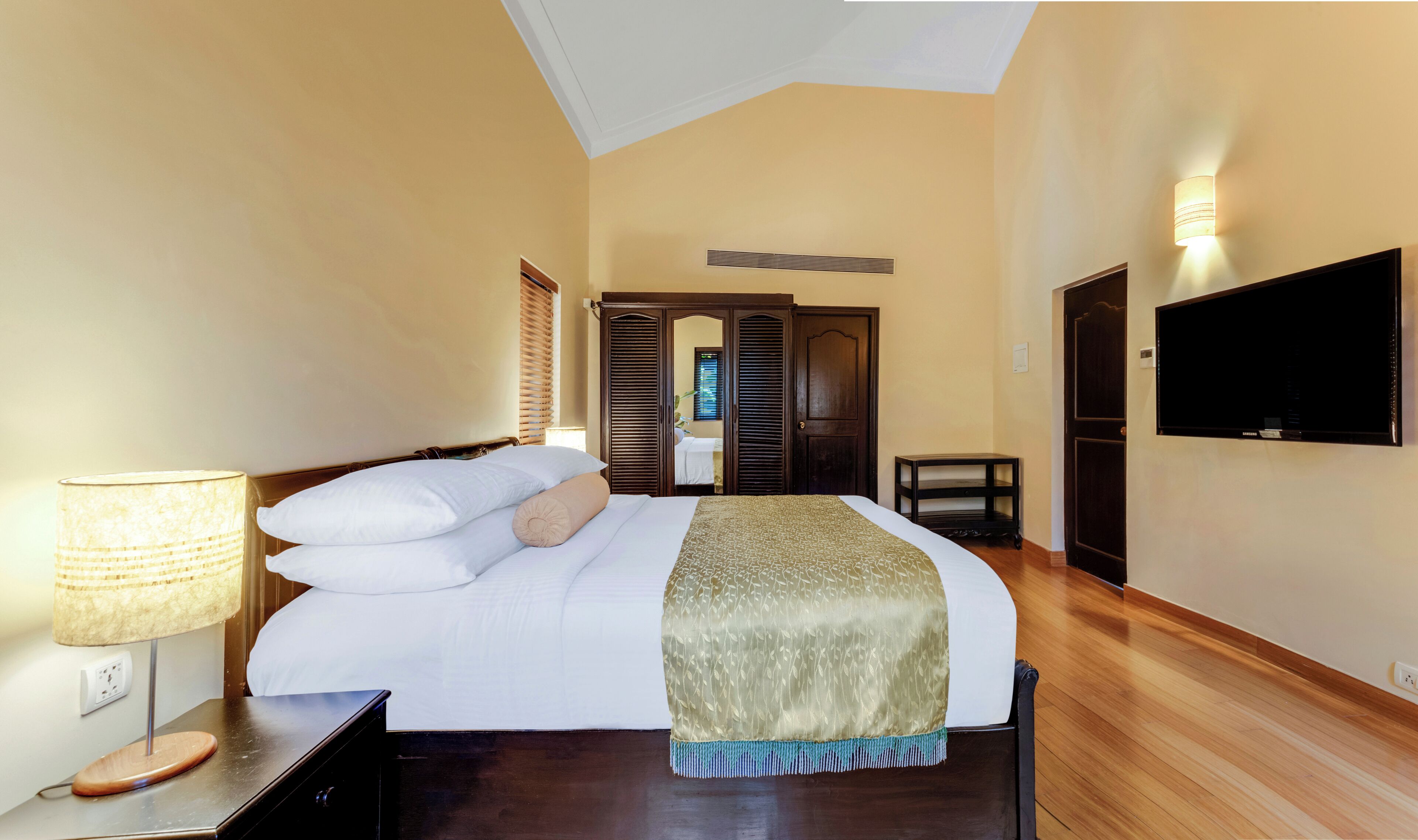 Quarto de hotel com cama, luminária de cabeceira, televisão, guarda-roupa e espelho.