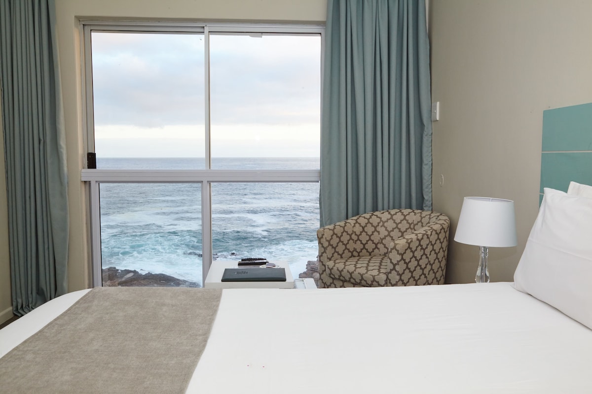 Quarto de hotel com uma janela grande, uma cama, uma cadeira e uma mesa de cabeceira.