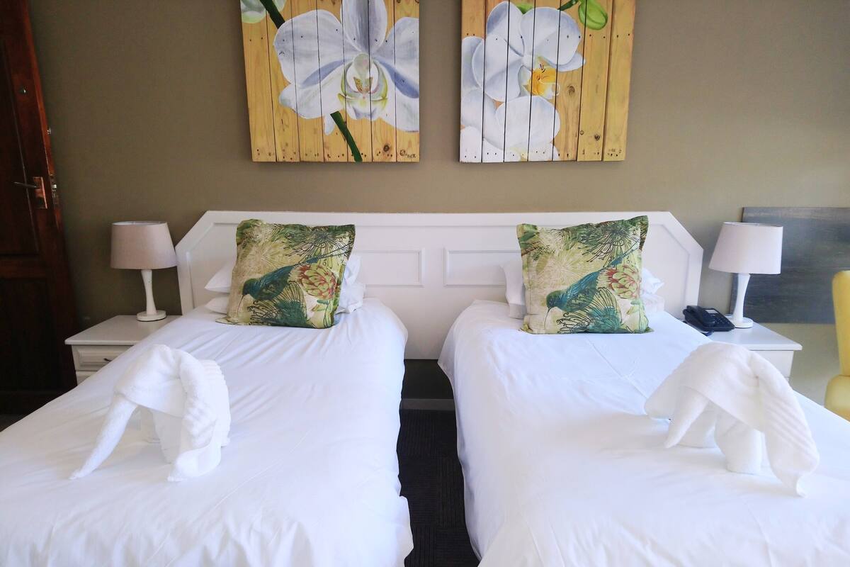 Quarto de hotel com duas camas, roupa de cama branca e quadros com motivos florais nas paredes.
