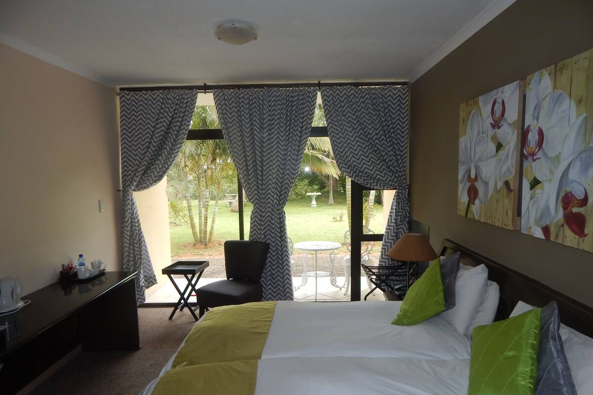 Quarto de hotel com cama, escrivaninha, cadeira e mesa. Há uma janela com cortinas e vista para o exterior.