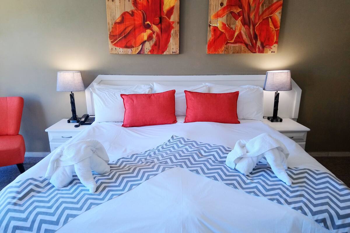 Quarto de hotel com uma cama, dois travesseiros vermelhos, uma colcha branca, um cobertor azul e branco com padrão listrado em chevron, duas luminárias fixadas na parede e uma cadeira vermelha.