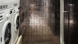 Uma lavanderia com máquinas de lavar e secar empilhadas, uma parede revestida de azulejos com textura e uma porta de metal.