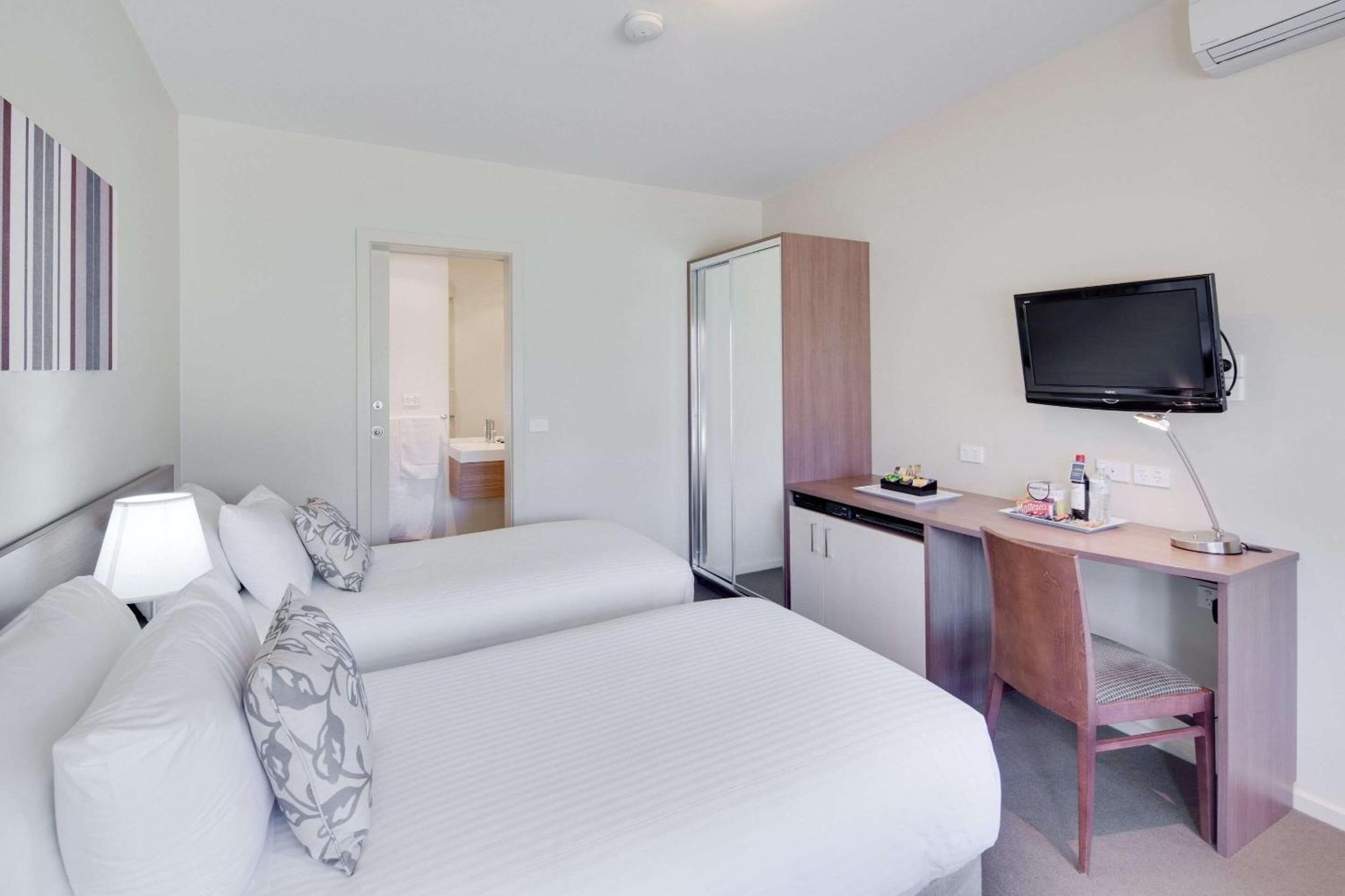 Quarto de hotel com duas camas, uma escrivaninha com cadeira, televisão e uma pequena cozinha.
