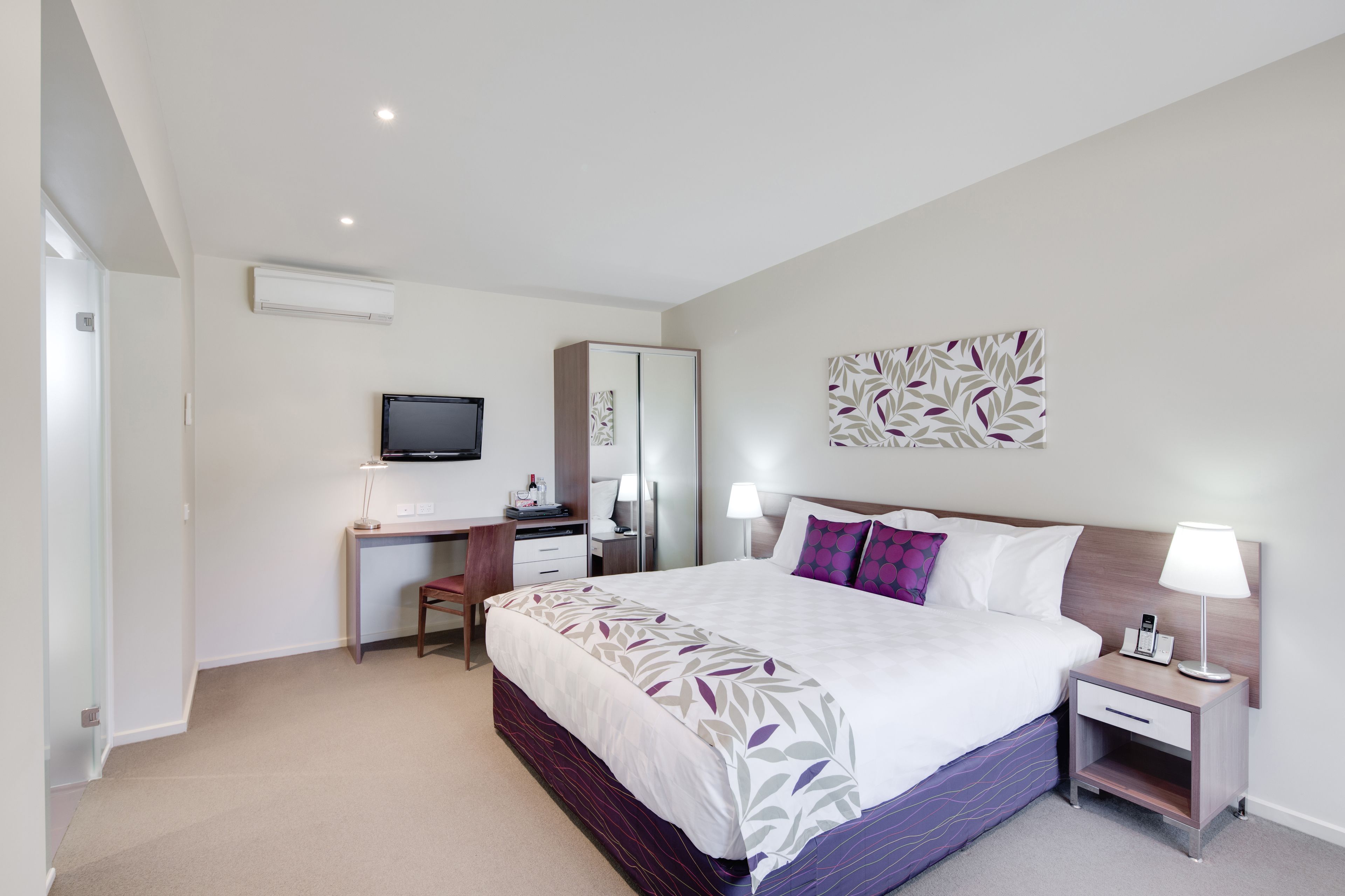 Quarto de hotel com cama, escrivaninha, cadeira, televisão, guarda-roupa e mesa de cabeceira com abajur e telefone.