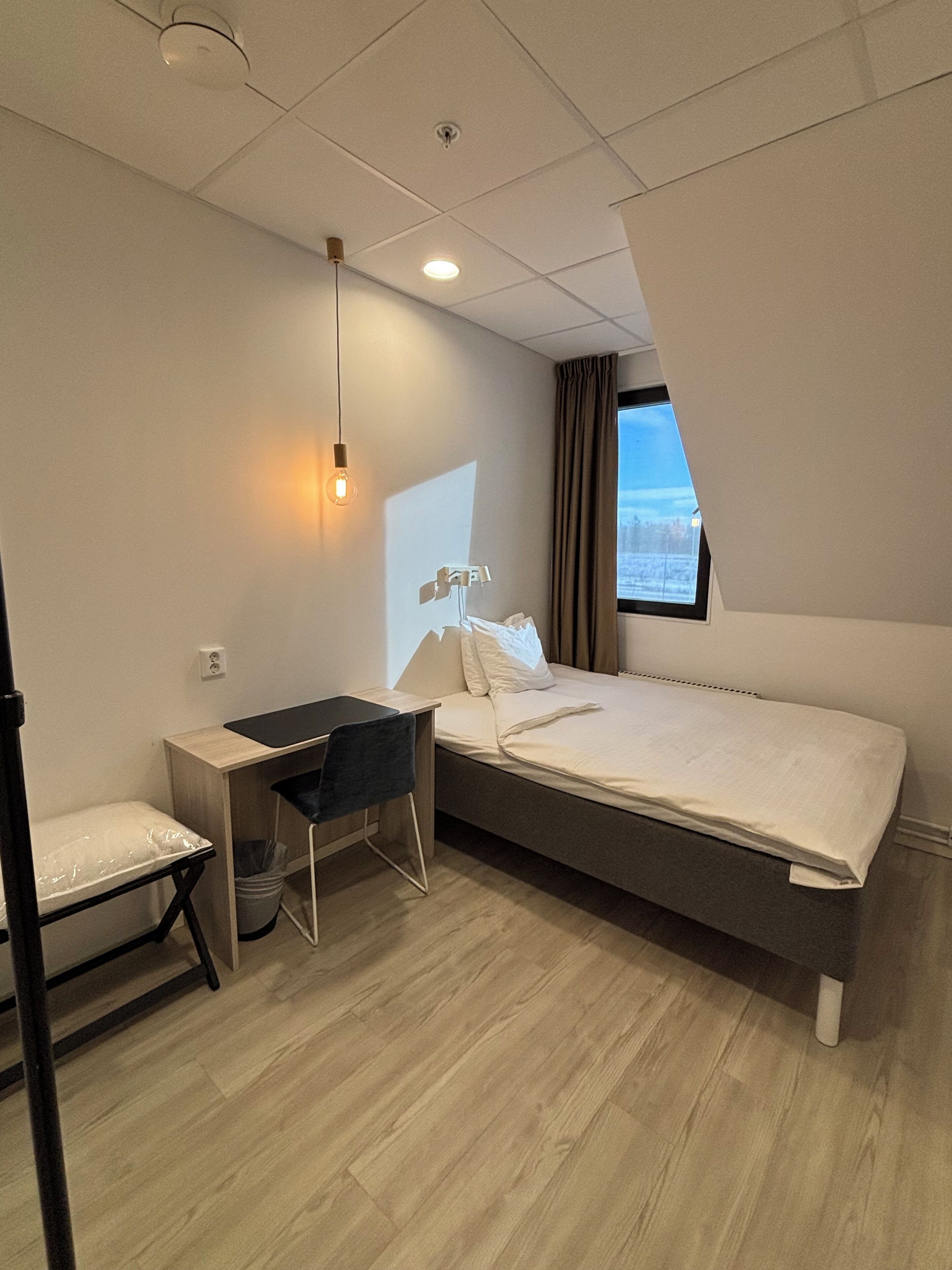 Quarto de hotel moderno com cama, escrivaninha, cadeira e janela com cortinas.