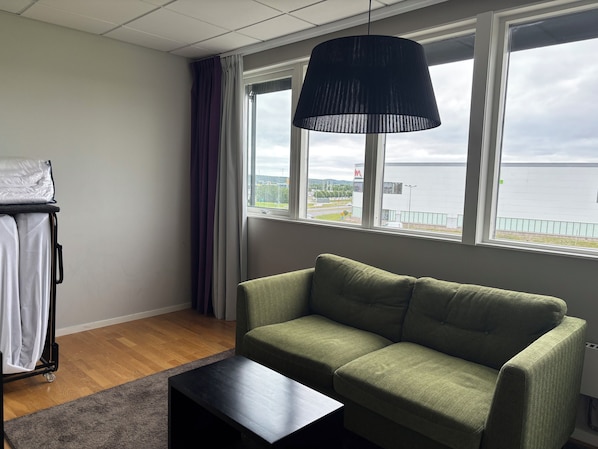 Quarto com sofá verde, mesa preta, janela com vista para um edifício e um corpo de água, e teto com padrão quadriculado.