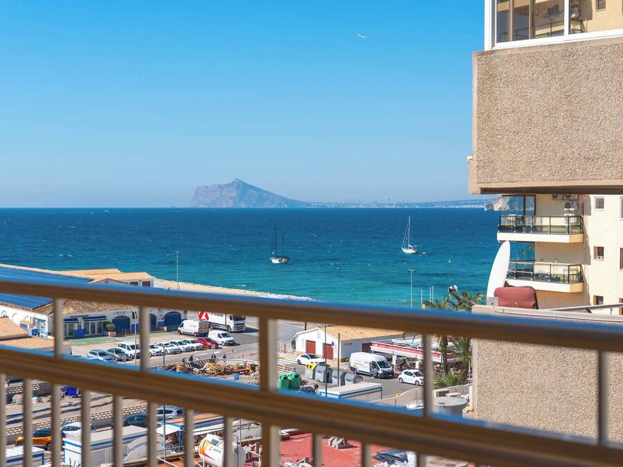 Vista costeira com montanha, barcos e um edifício com varandas.