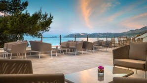 Um terraço com mesas e cadeiras ao ar livre, uma mesa de vidro e vista para o mar e montanhas.