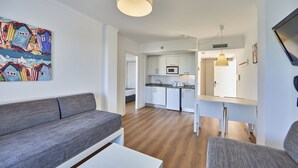 Uma sala de estar moderna com um sofá cinzento, uma mesa de centro branca e um espaço de jantar com uma mesa e cadeiras brancas. Cozinha ao fundo com armários brancos e um micro-ondas.