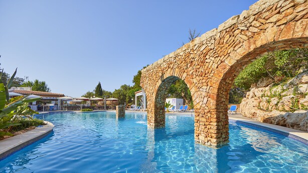 Uma ponte de pedra em arco sobre uma piscina, com espreguiçadeiras e um edifício ao fundo.