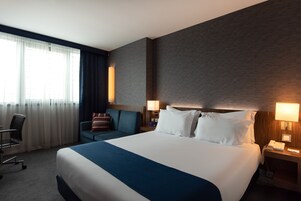 Quarto de hotel com uma cama grande, uma escrivaninha, uma cadeira e uma janela com cortinas.