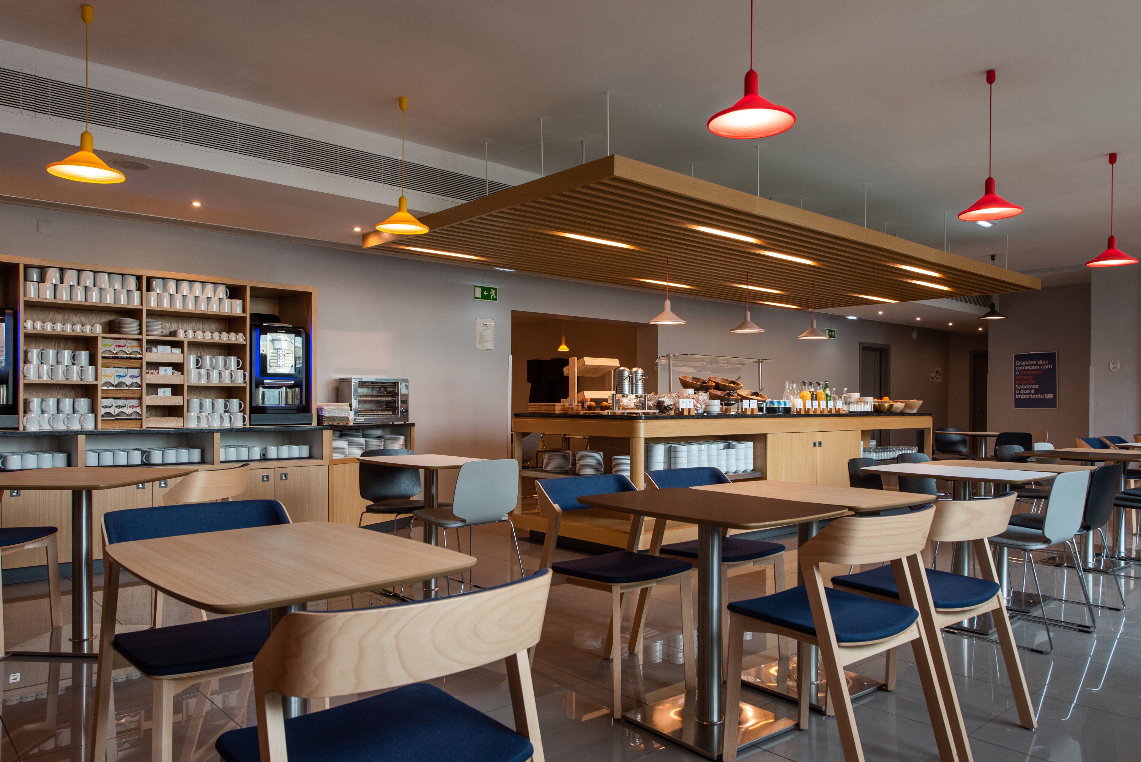 Um restaurante moderno com mesas de madeira e cadeiras azuis, um balcão com diversas bebidas, e uma exposição de canecas e pratos.