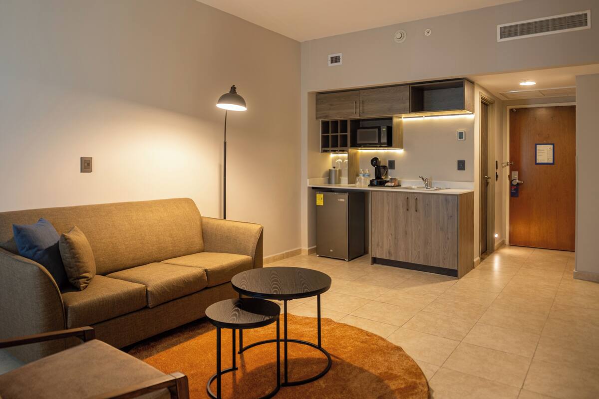 Uma sala de estar moderna com um sofá, uma pequena mesa redonda e um abajur. Próximo à sala de estar, encontra-se uma cozinha compacta com micro-ondas, pia e armários.