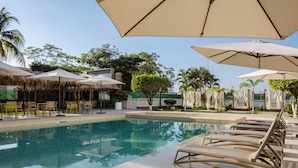 Uma piscina com espreguiçadeiras e guarda-sóis, cercada por árvores e uma construção com teto de palha.
