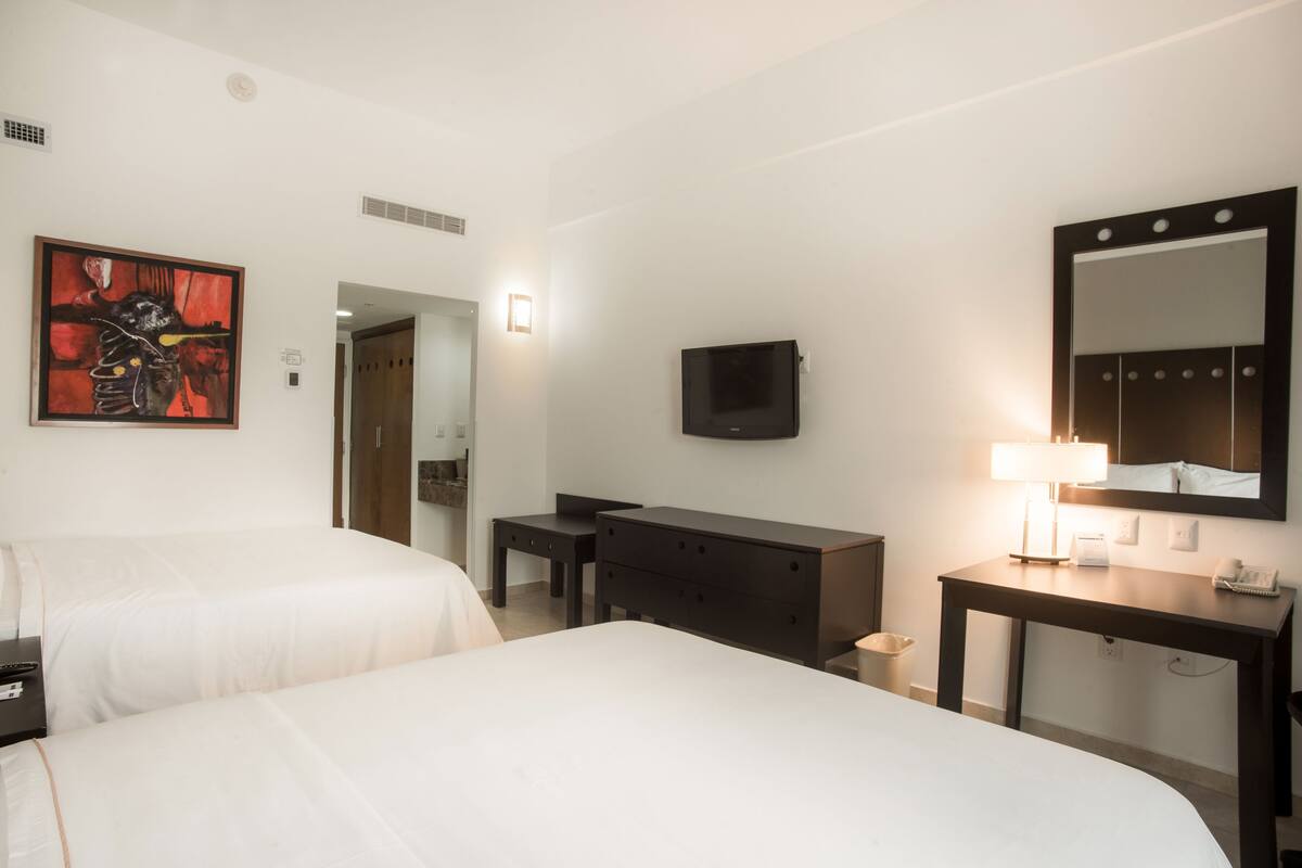 Quarto de hotel com duas camas, uma escrivaninha, cômoda, televisão, espelho e um quadro na parede.