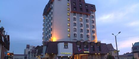 Um hotel de vários andares com uma placa de 'Continental' bem visível, um estacionamento com vários carros e um céu claro.