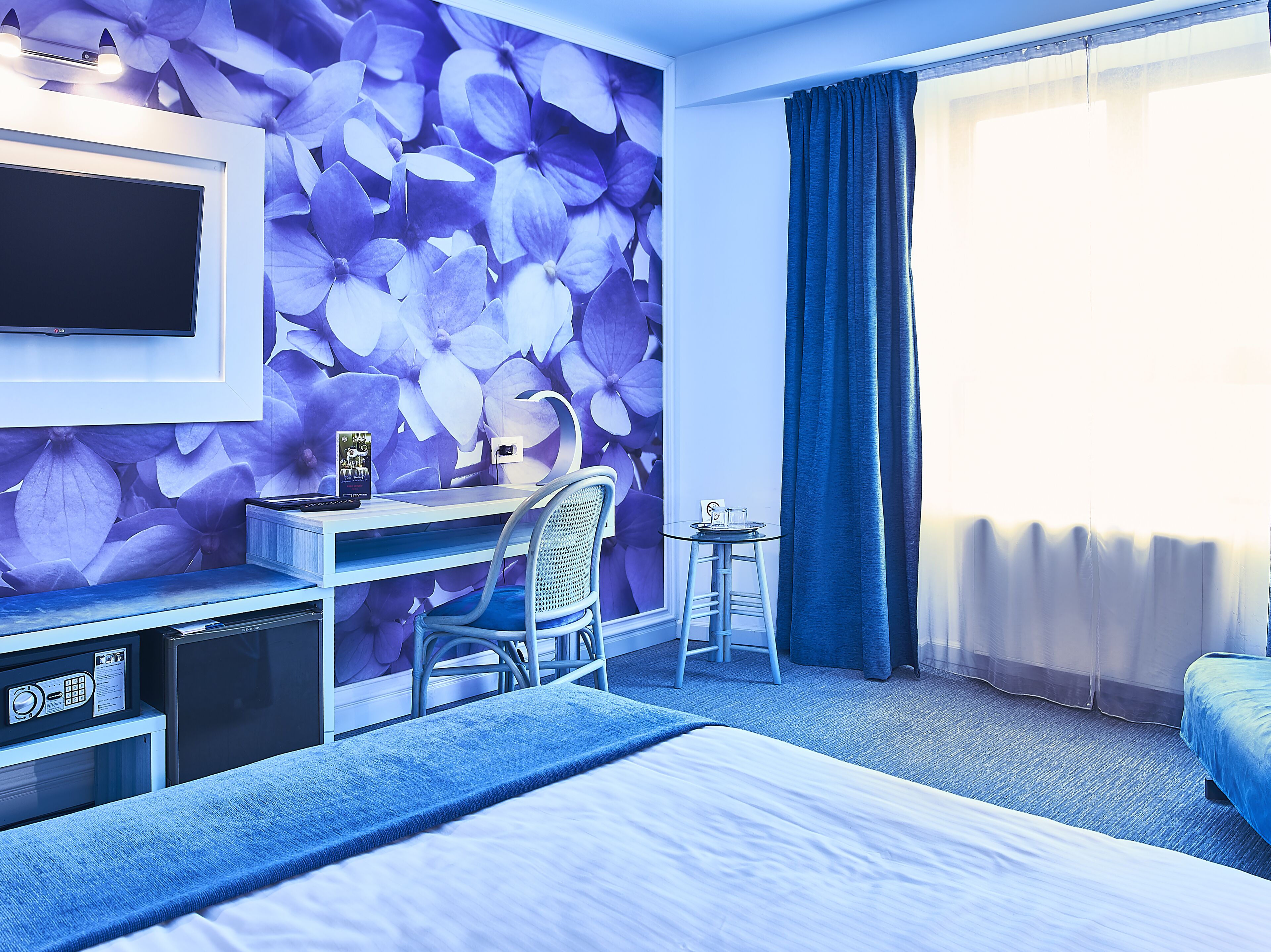 Quarto de hotel com cama, escrivaninha, cadeira, televisão e janela com cortinas.