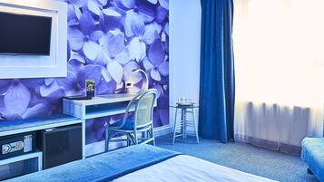 Quarto de hotel com cama, escrivaninha, cadeira, televisão e janela com cortinas.