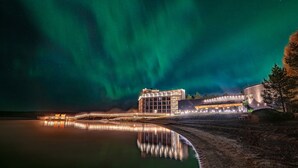 Um edifício moderno com fachada de vidro, localizado à beira de águas tranquilas que refletem luzes, sob um céu noturno com a aurora boreal.