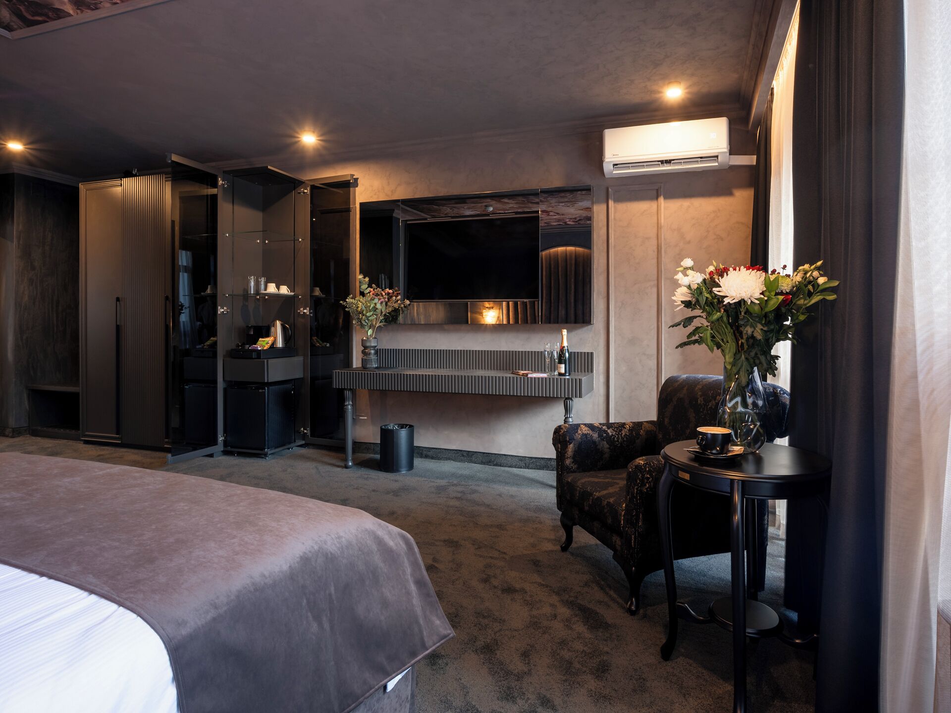 Quarto de hotel com cama, área de estar com poltrona e mesa, televisão de tela plana na parede e uma pequena lixeira.