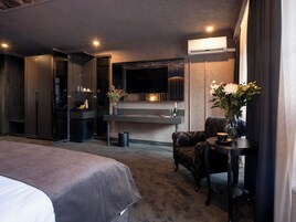 Quarto de hotel com cama, área de estar com poltrona e mesa, televisão de tela plana na parede e uma pequena lixeira.