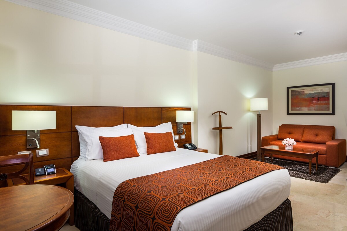 Quarto de hotel com cama grande, duas mesas de cabeceira, escrivaninha, cadeira e sofá.