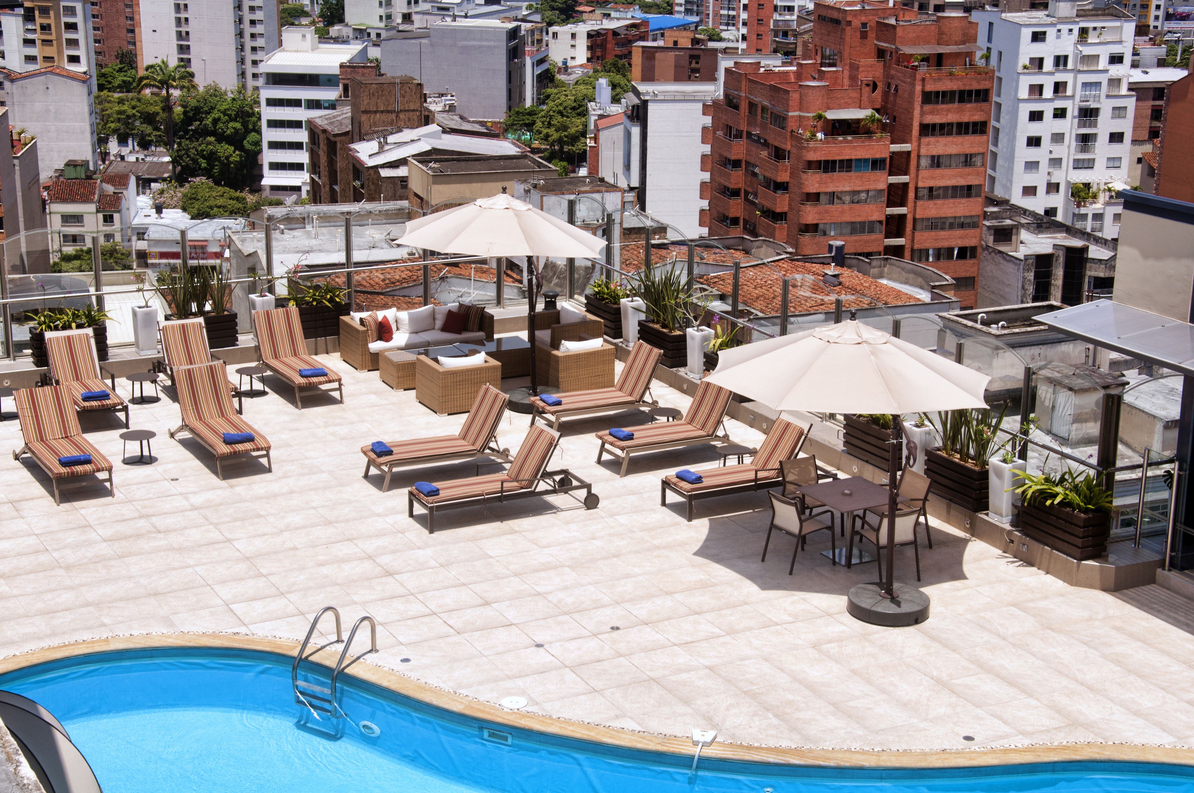 Uma área de piscina no terraço com espreguiçadeiras e guarda-sóis, rodeada por edifícios da cidade.