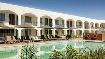 Uma área à beira da piscina com espreguiçadeiras e guarda-sóis, um céu azul límpido, e um edifício com janelas em arco e varandas.