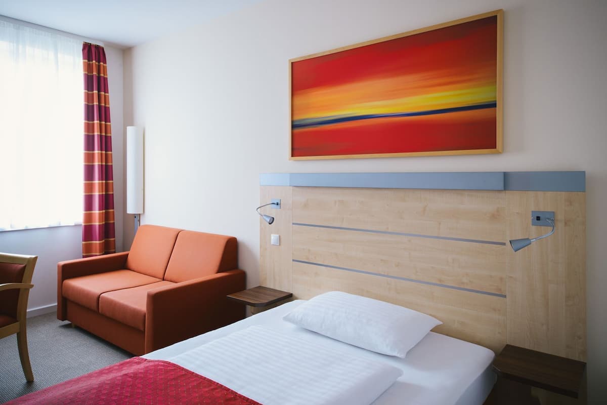 Quarto de hotel com cama, cadeira, escrivaninha, pintura e janela com cortinas.
