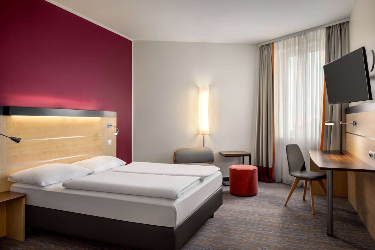 Quarto de hotel com cama, escrivaninha, cadeira, televisão, abajur e um banquinho vermelho.